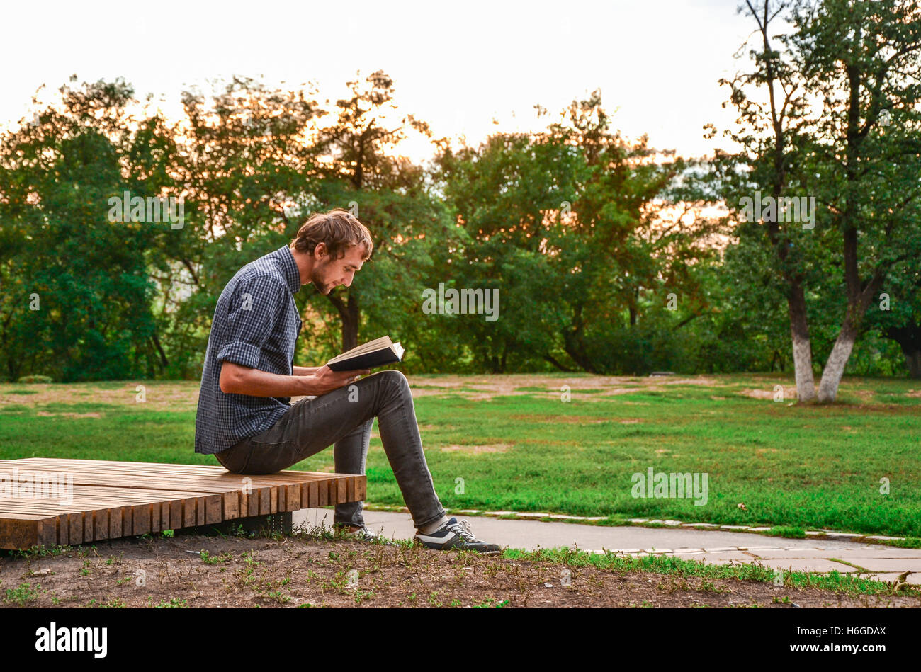 Guy sitting on a bench in the park reading book Stock Photo - Alamy