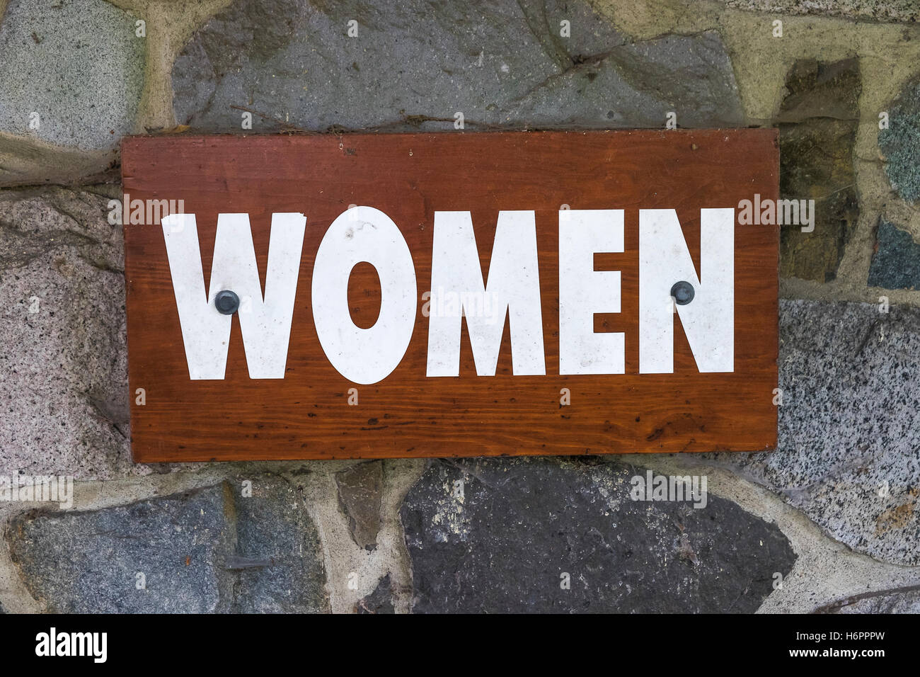 A Women sign painted on wood hangs on a stone wall outside a washroom ...