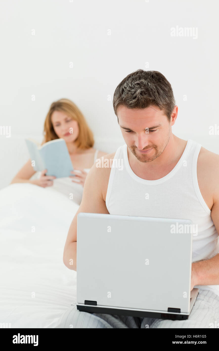 Man is on his laptop while his wife is reading a book in their bed ...