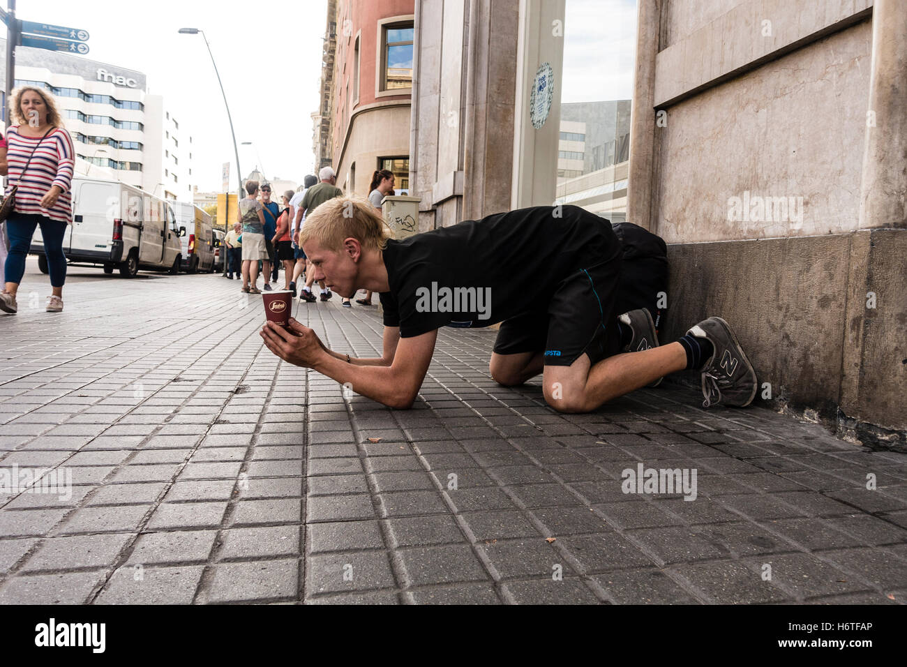 A young male beggar kneels on the sidewalk holding his cup as he begs ...