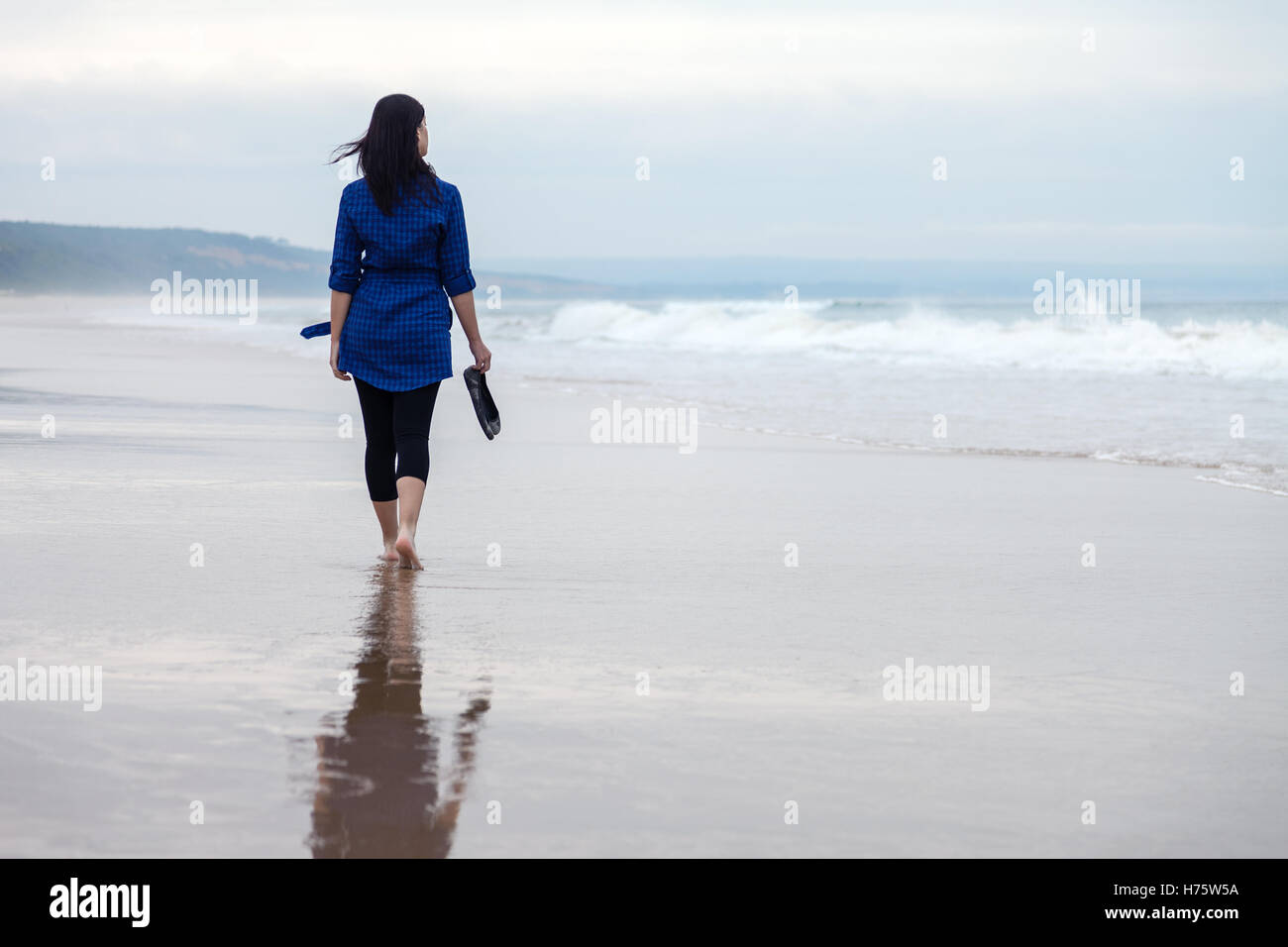 Young woman walking alone in a deserted beach reflected on the wet sand ...