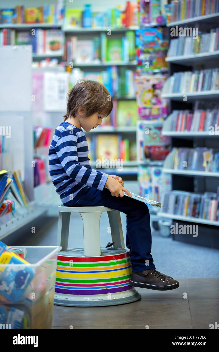 Adorable little child, boy, sitting in a book store, reading books ...
