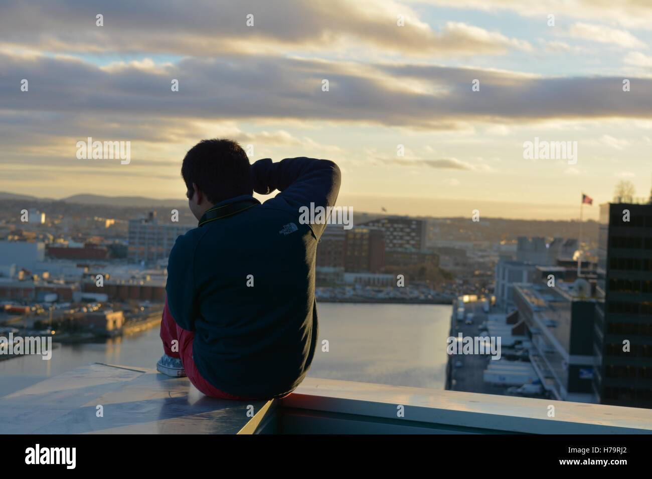 A man sitting on a ledge of a skyscraper overlooking a city at sunset ...