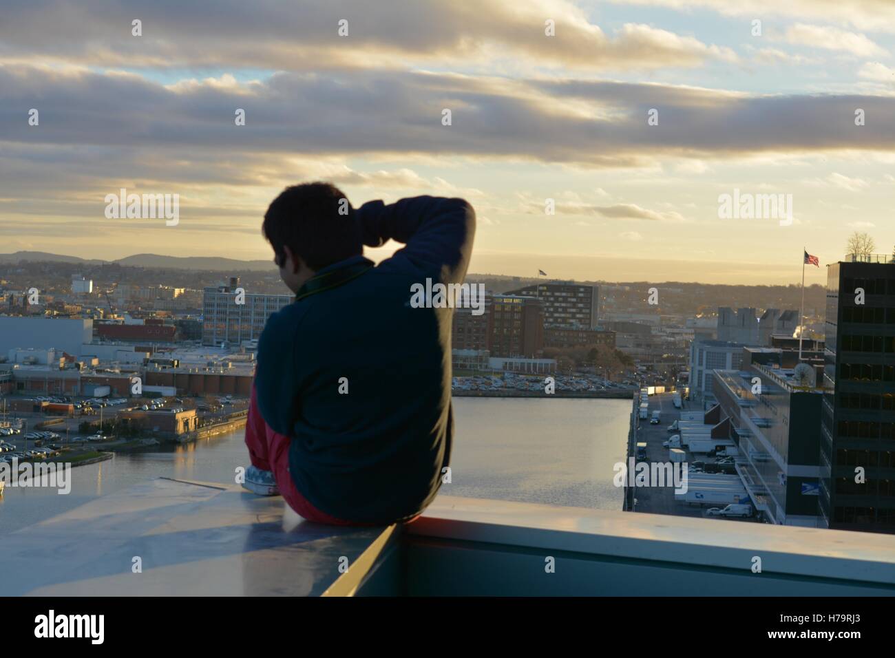 A man sitting on a ledge of a skyscraper overlooking a city at sunset ...