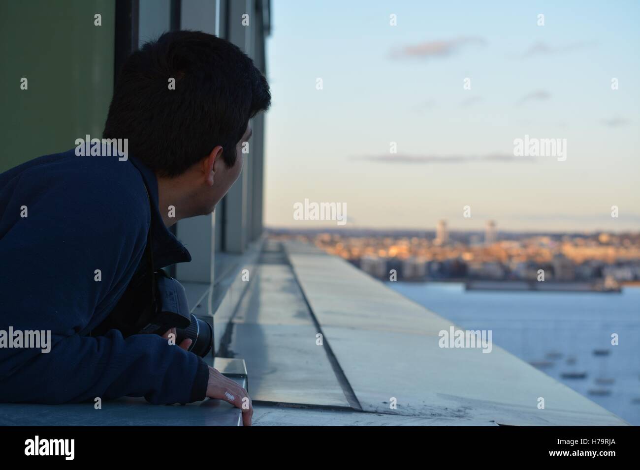 A man looking over a ledge of a skyscraper overlooking a city at sunset ...