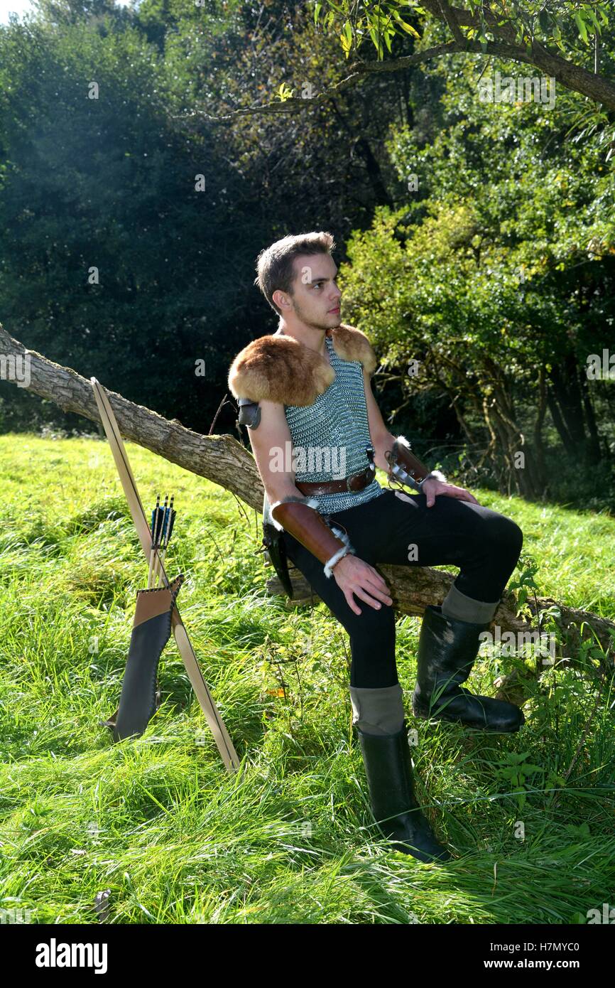 Young Medieval dressed man, sits on branch, arms on legs and looks ...