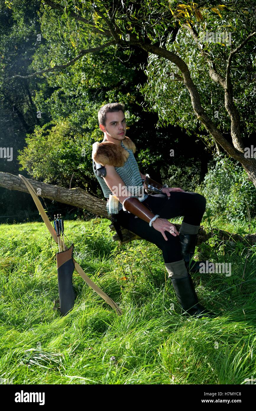 Young Medieval dressed man, sits on branch, arms on legs and looks ...