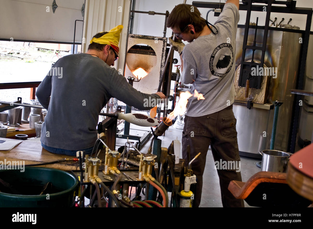 Two men are working in a shop, doing glass blowing with a torch and ...