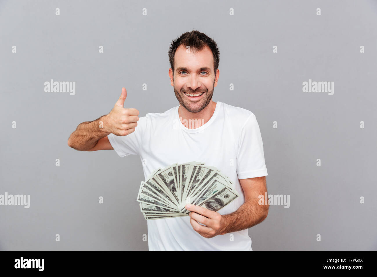 Cheerful successful young man holding money and showing thumbs up over ...