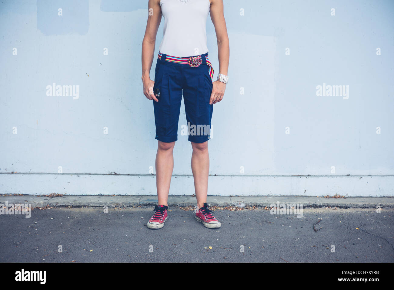 A stylish young woman wearing shorts is standing by a blue wall outside ...