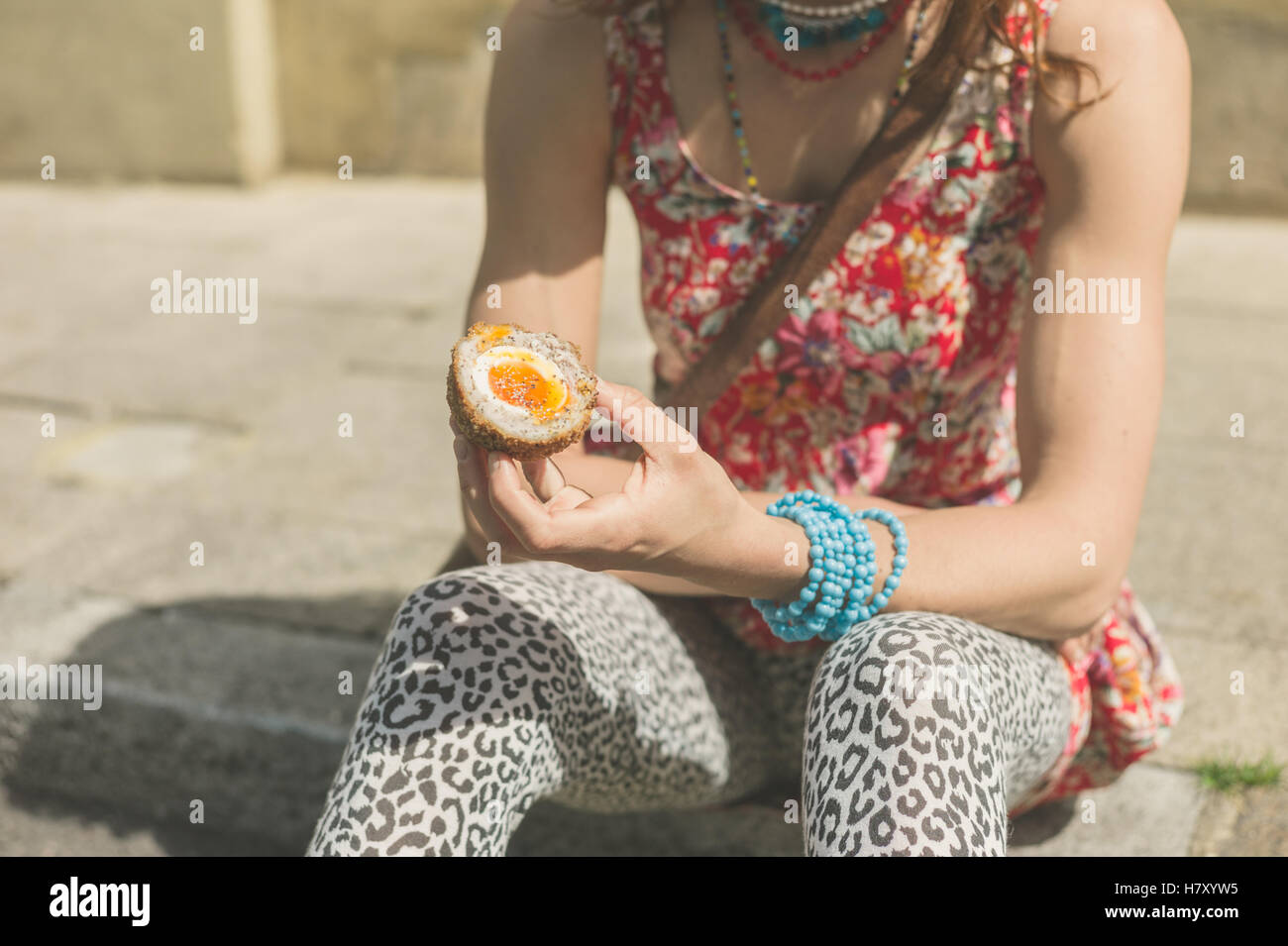 A young woman is sitting on the curb on the street and is eating a ...