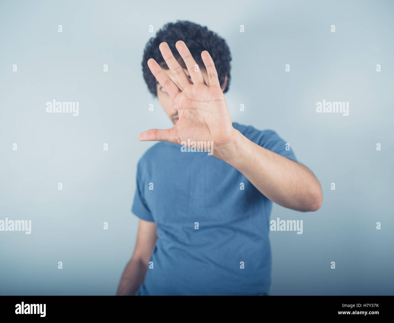 A young man wearing blue is raising his hand to cover and hide his face ...