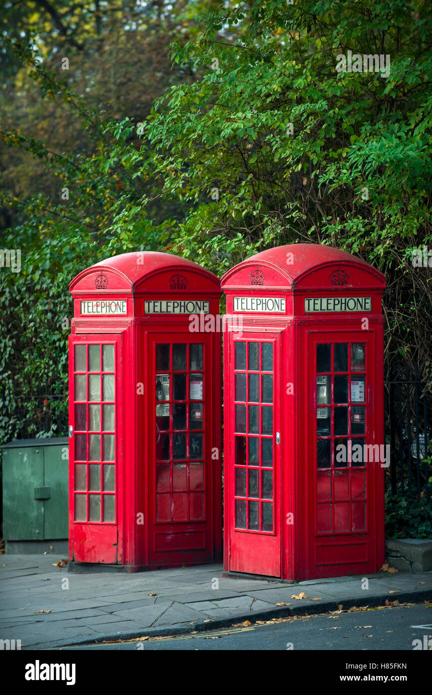 Pair of classic red telephone booths stand under greenery at the edge ...