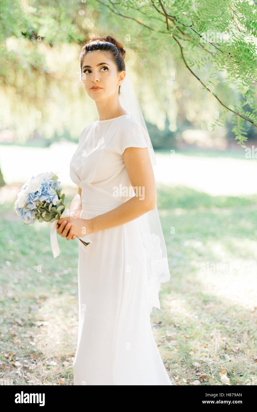 Thoughtful bride in simple white dress stands under green tree Stock ...