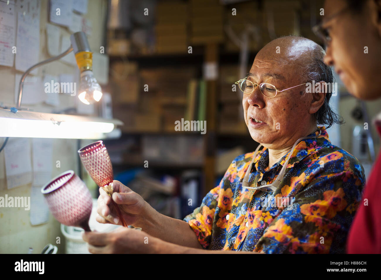 Two people, a father and son at work in a glass maker's studio workshop, inspecting a red cut ...