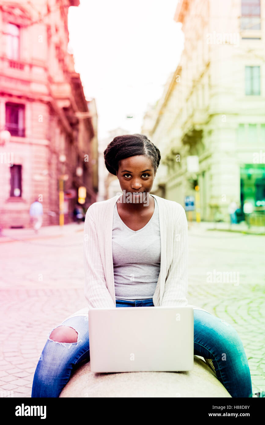 Young black woman using computer outdoor in the city sitting on a bench ...