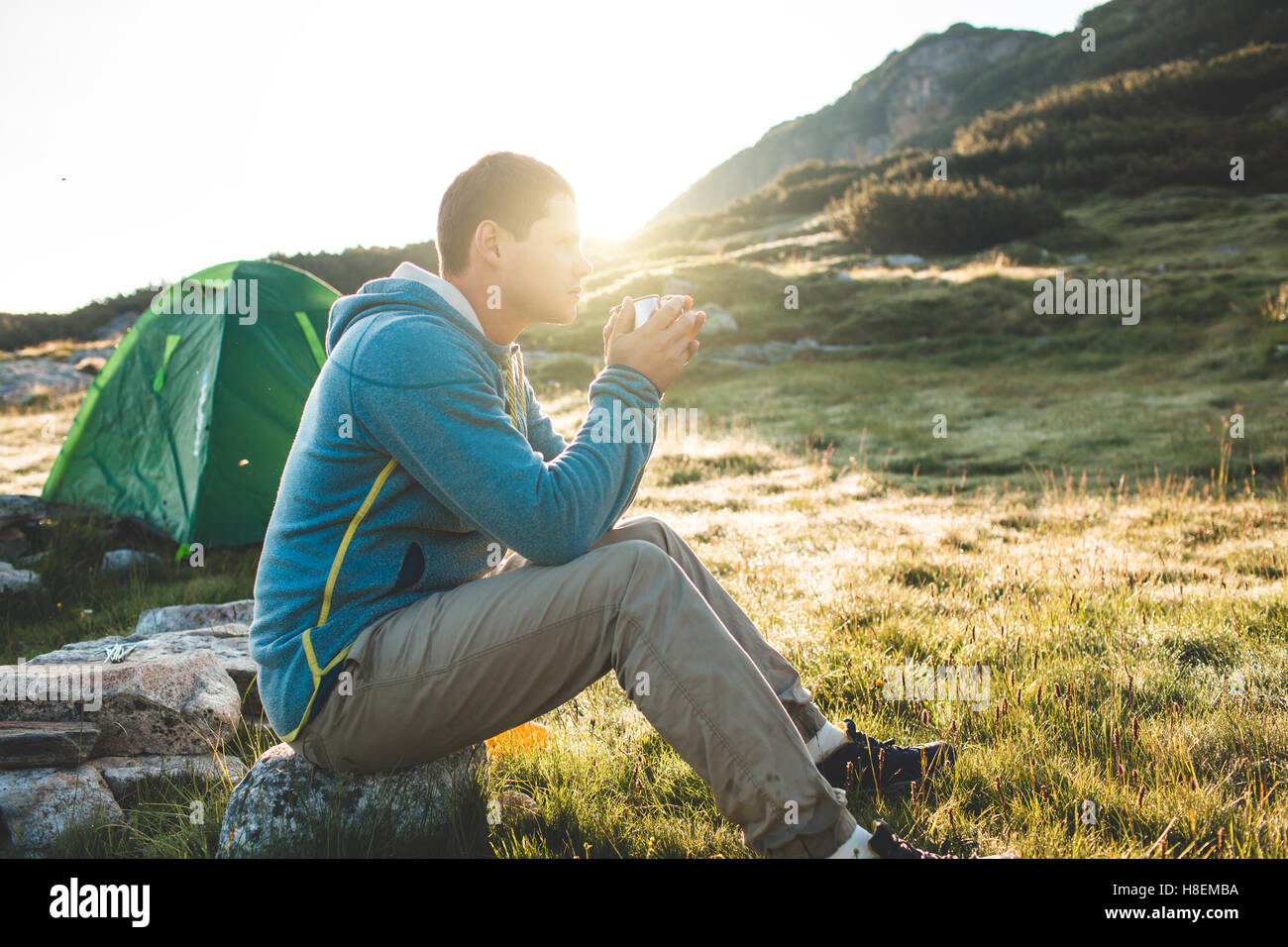 Young man drinking tea at sunrise in mountains. Mountain camping ...