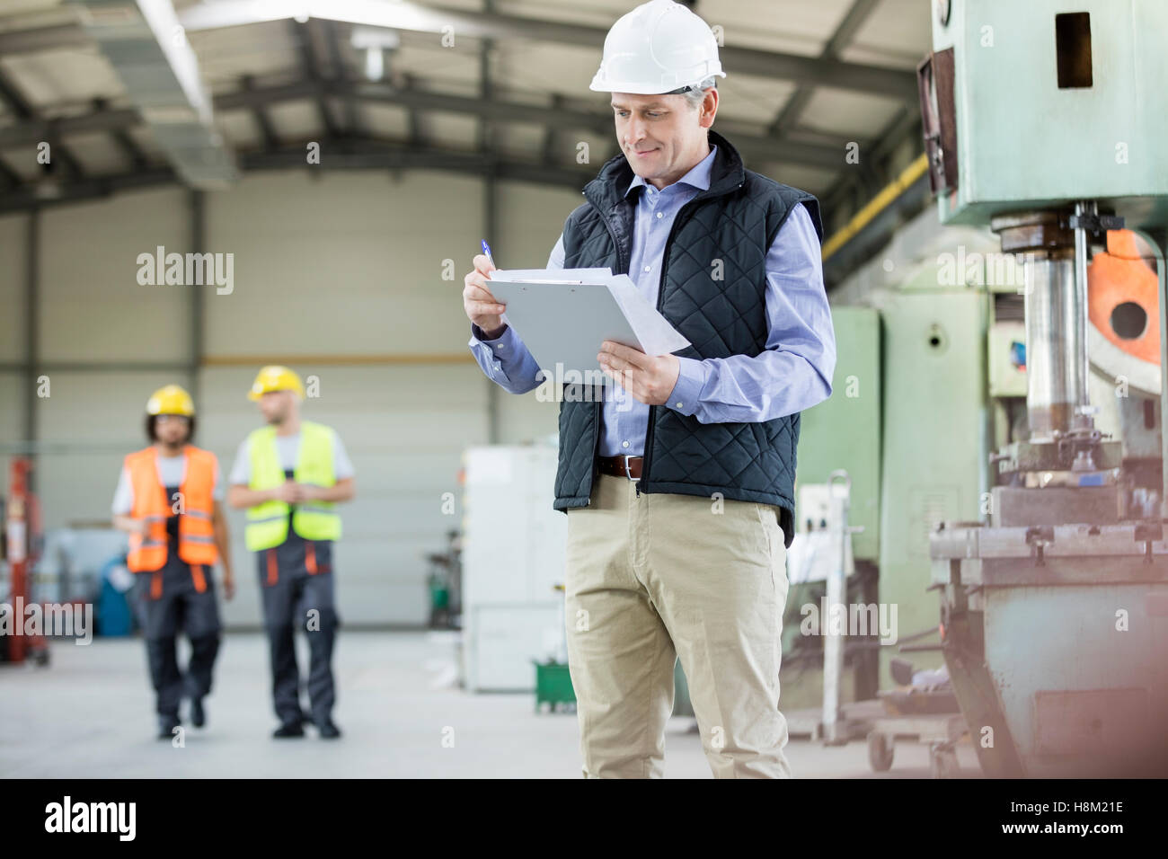 Mature male inspector writing on clipboard while workers in background ...