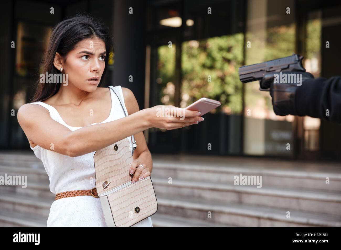 Frightened young woman giving mobile phone to robber in gloves ...