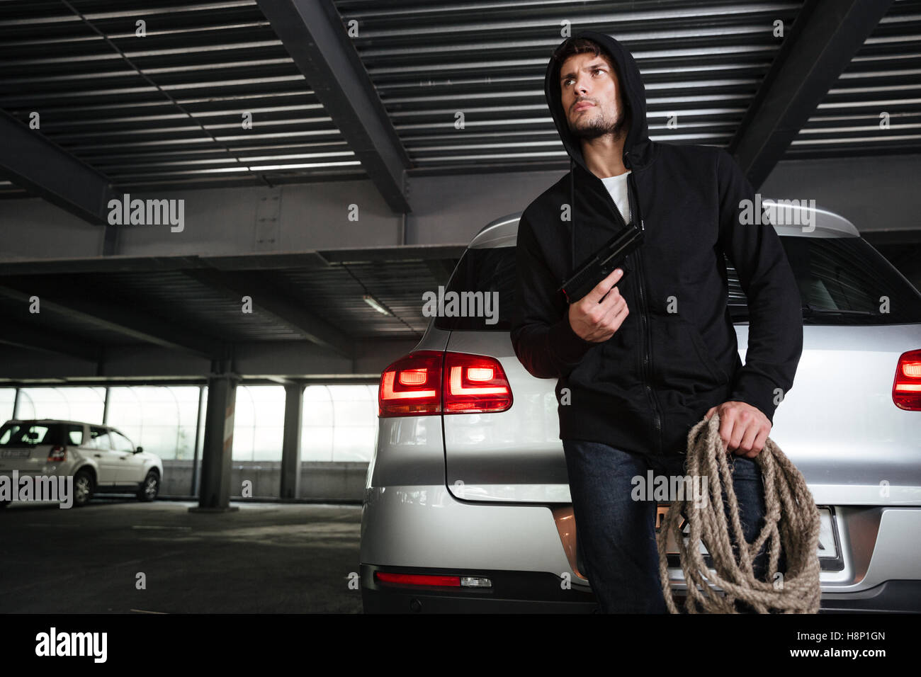 Dangerous criminal young man with gun and rope standing on car parking ...