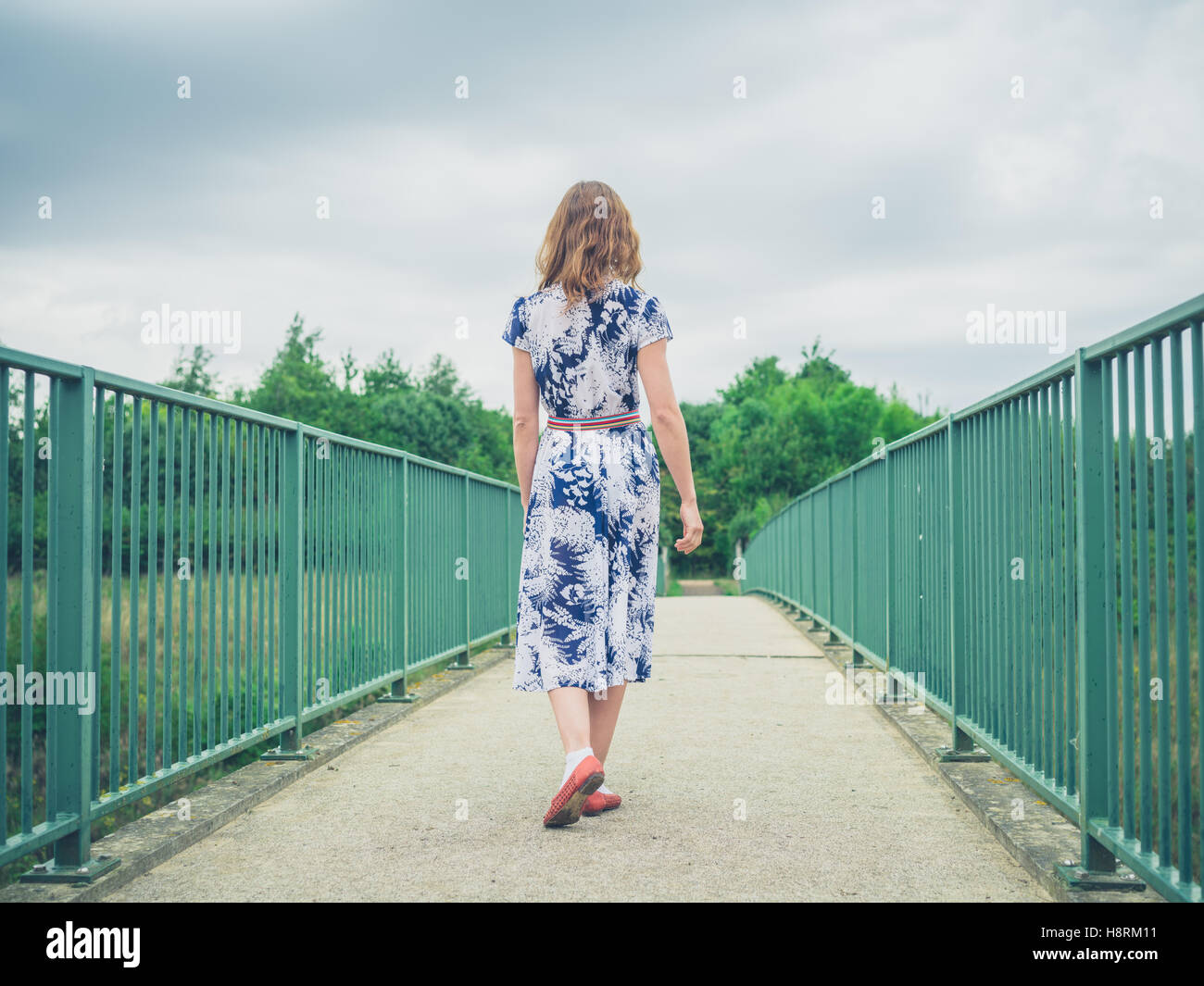 A young woman wearing a dress is crossing a footbridge in a rural ...