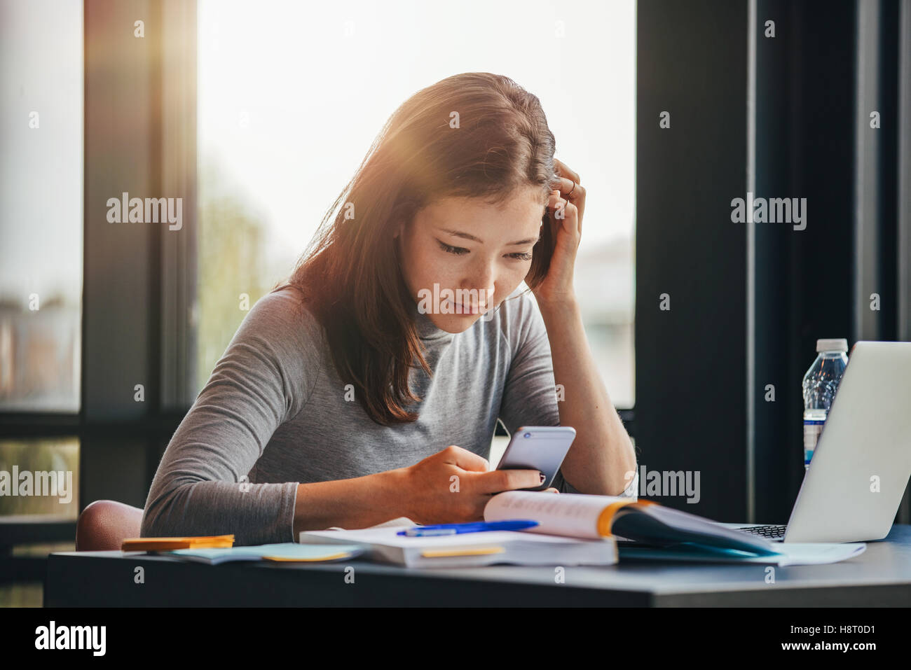 Shot of young woman sitting at library table reading text message on ...