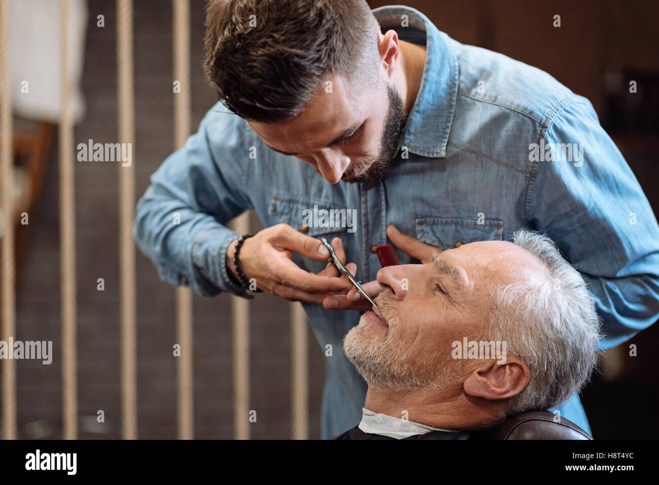 Handsome young barber cutting mustache of senior client with scissors ...