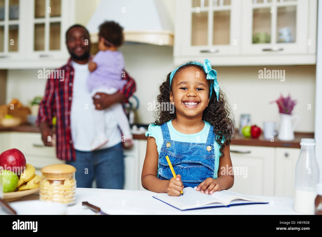Happy young girl looking at camera while doing homework in the kitchen ...