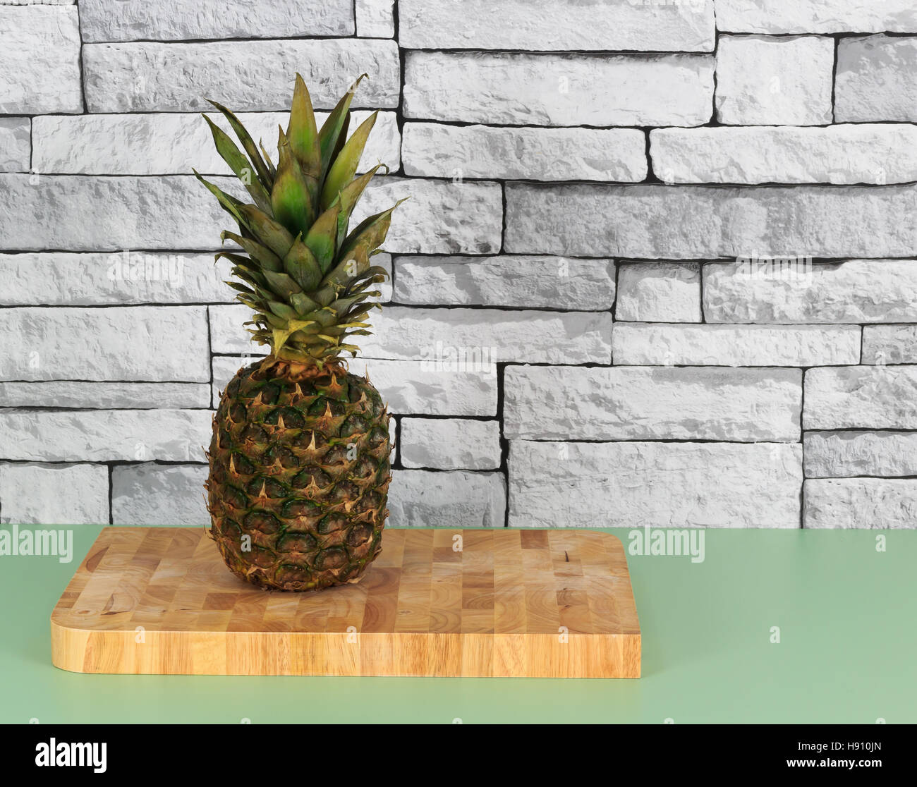Pineapple on wooden chopping board with old brick wall effect background and light green worktop ...