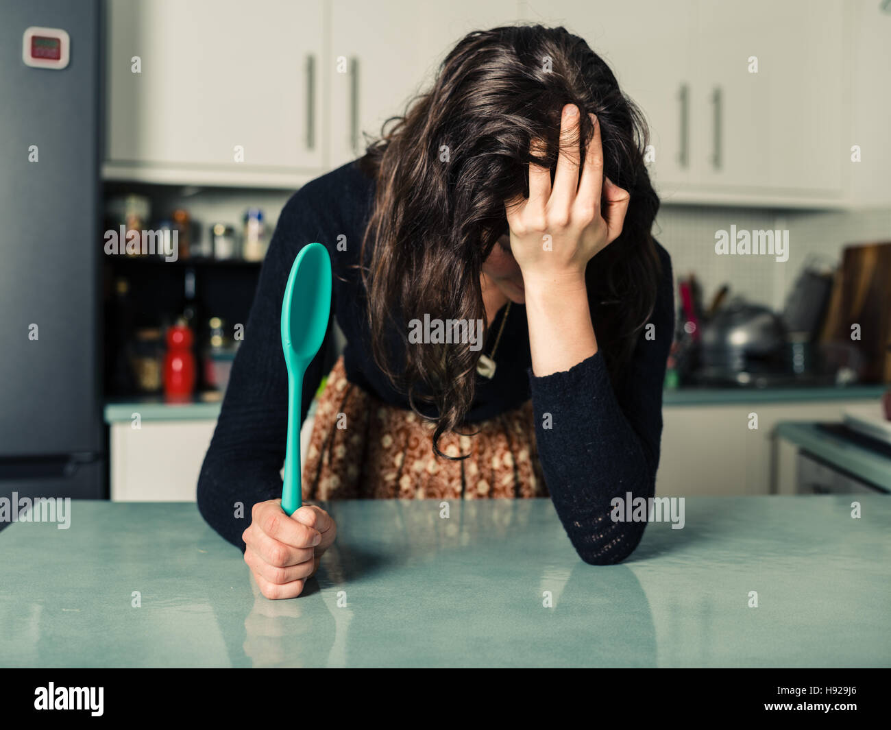 A sad young woman is standing in a kitchen with a spoon in her hand ...