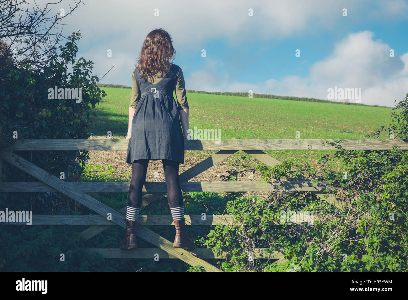 A young woman is standing on a gate in the countryside and is looking ...