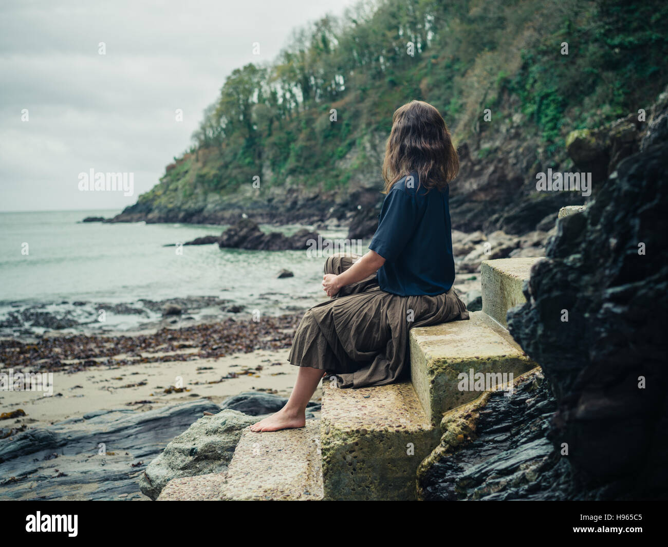 A young barefoot woman is sitting on some stairs outside by some rocks ...