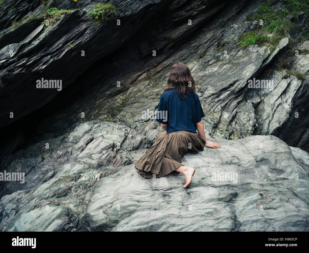 A young barefoot woman wearing a skirt is sitting on a rock outside a ...