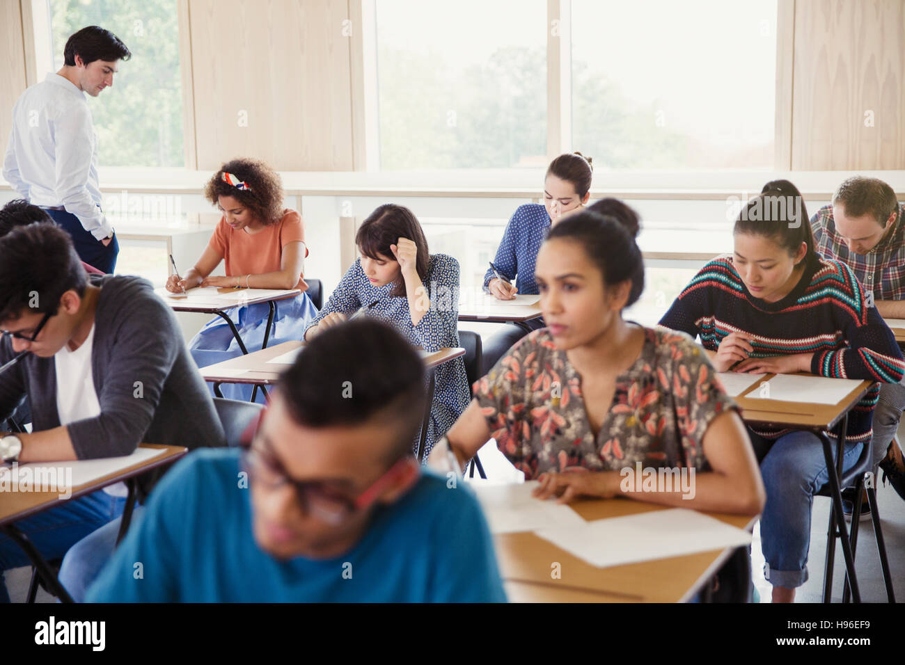 Professor watching college students taking test at desks in classroom ...