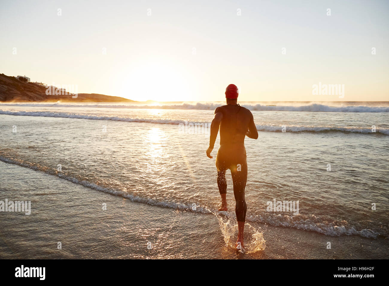 Male triathlete swimmer in wet suit running into ocean surf at sunrise ...