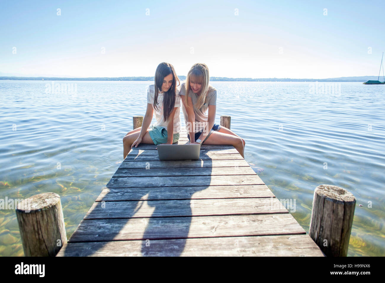 Two young women sit on pier by lake using laptop Stock Photo - Alamy