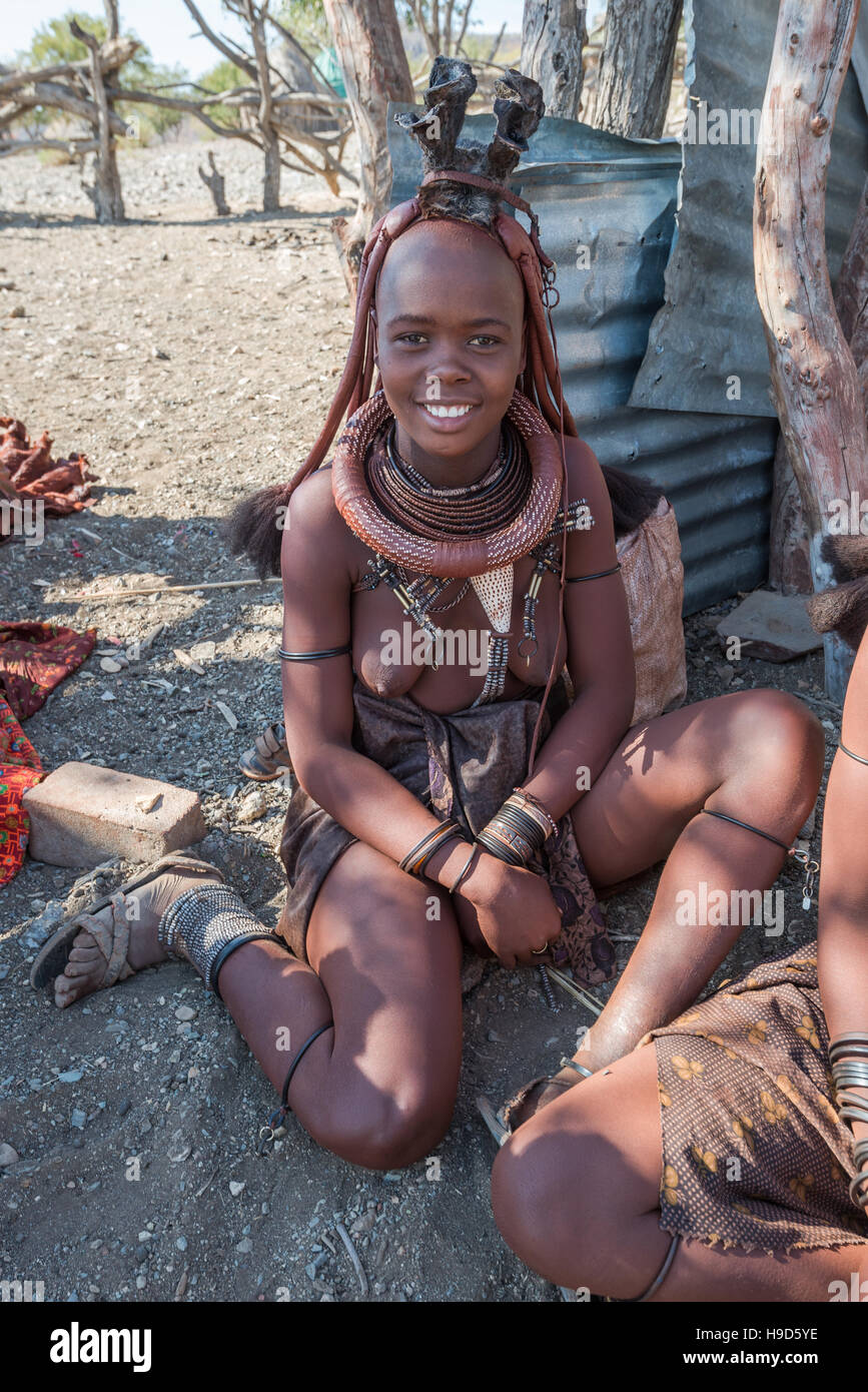 A young girl in traditional clothes and decoration from Himba people is sitting and trading at ...