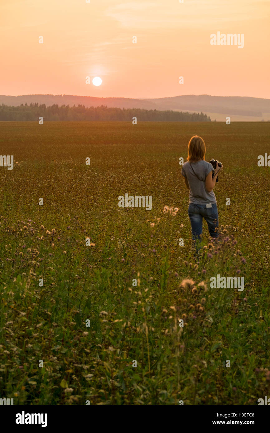 Caucasian woman walking in field at sunset with camera Stock Photo - Alamy