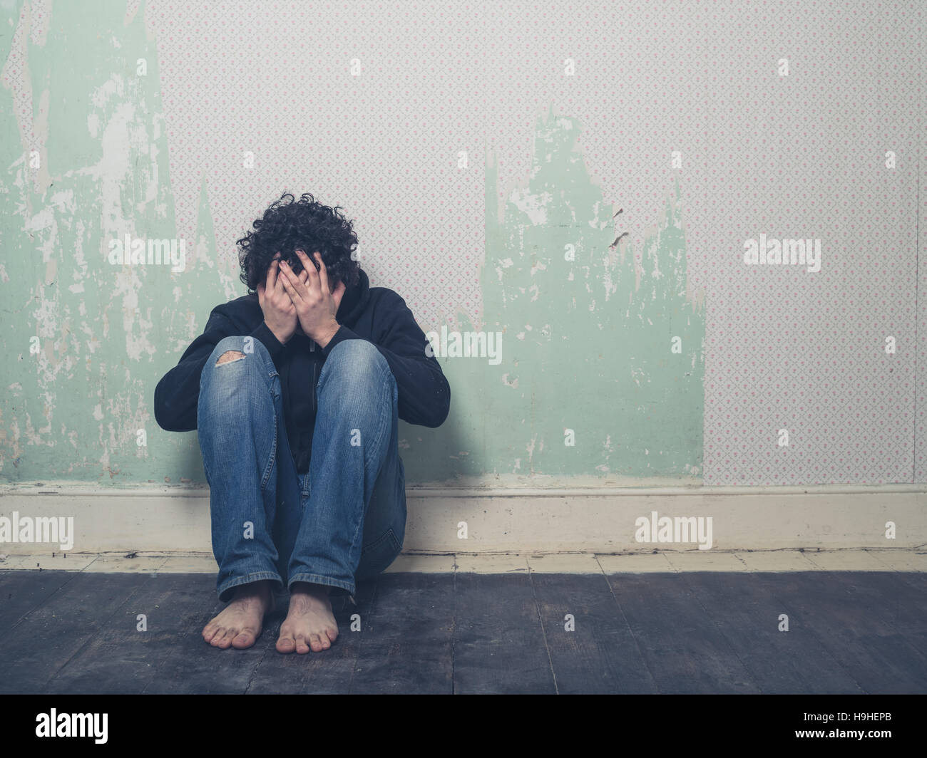 A sad and depressed young man is sitting on the floor in an empty room ...