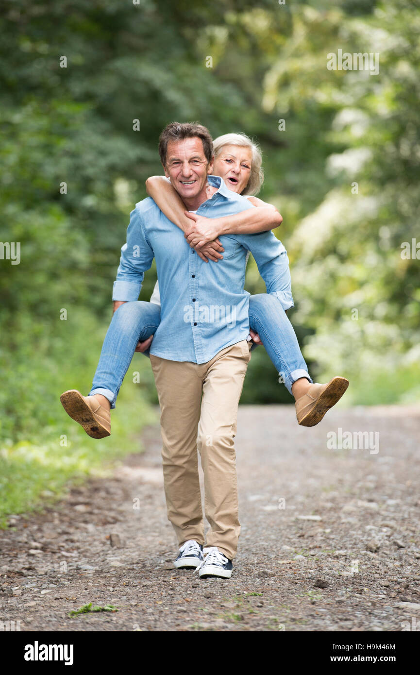 Smiling senior man giving his wife a piggyback ride on forest track ...