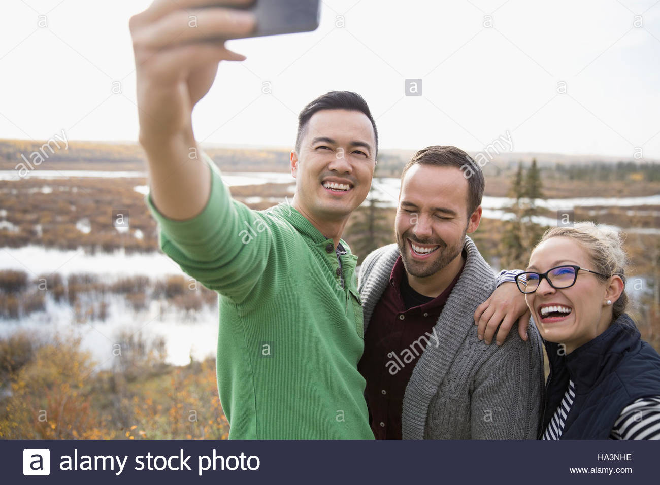 Smiling friends with camera phone taking selfie at autumn lakeside ...