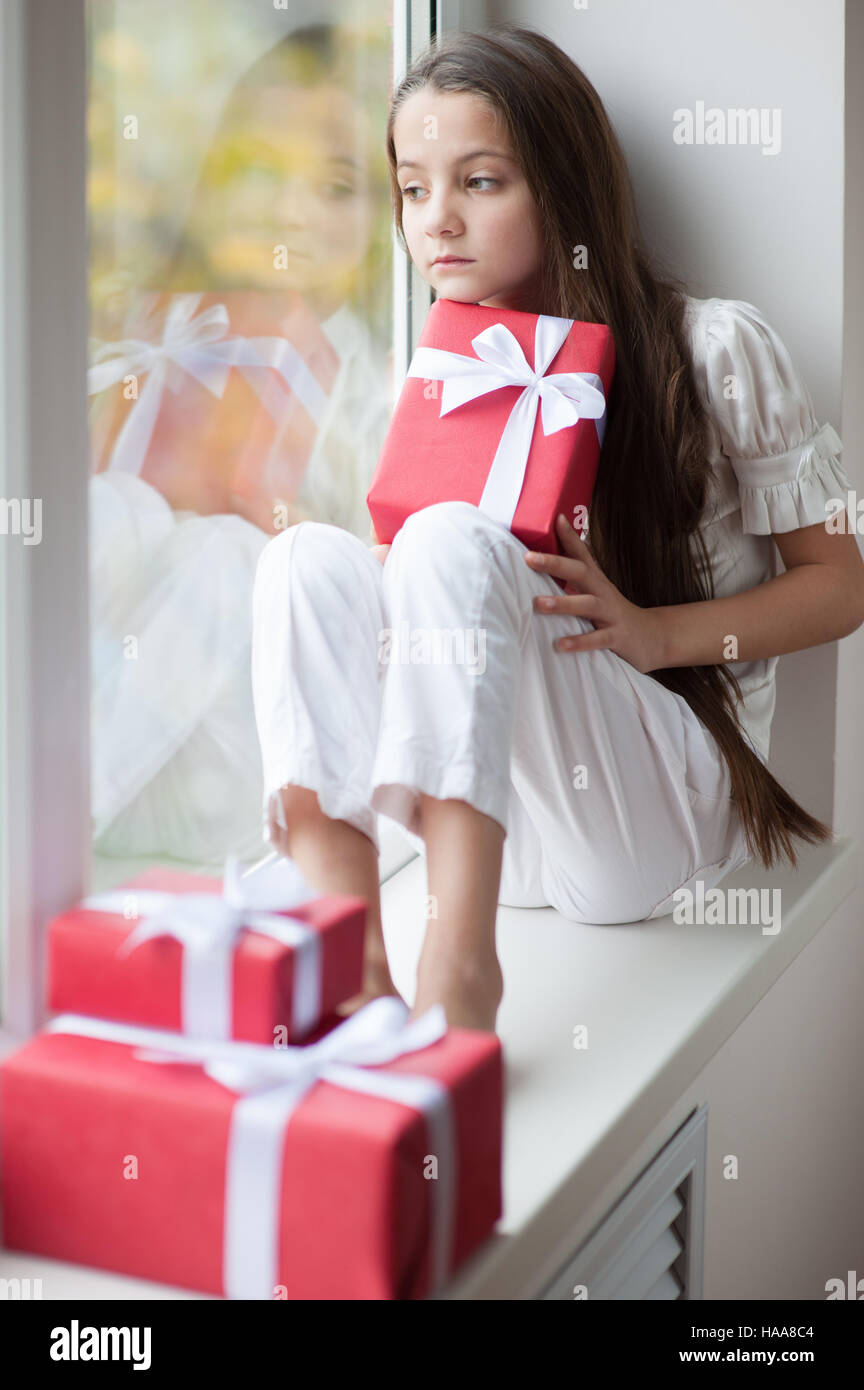 adorable little girl sitting by the window holding a box with gift near ...