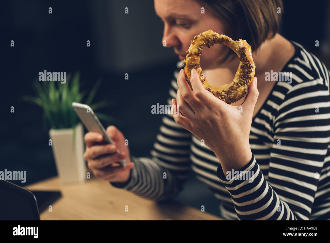Woman eating sesame bagel and using mobile phone in office while ...