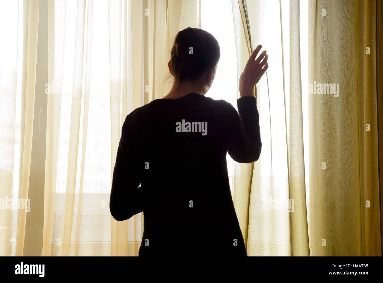 A woman in black moves the curtain aside to look out trough the window ...