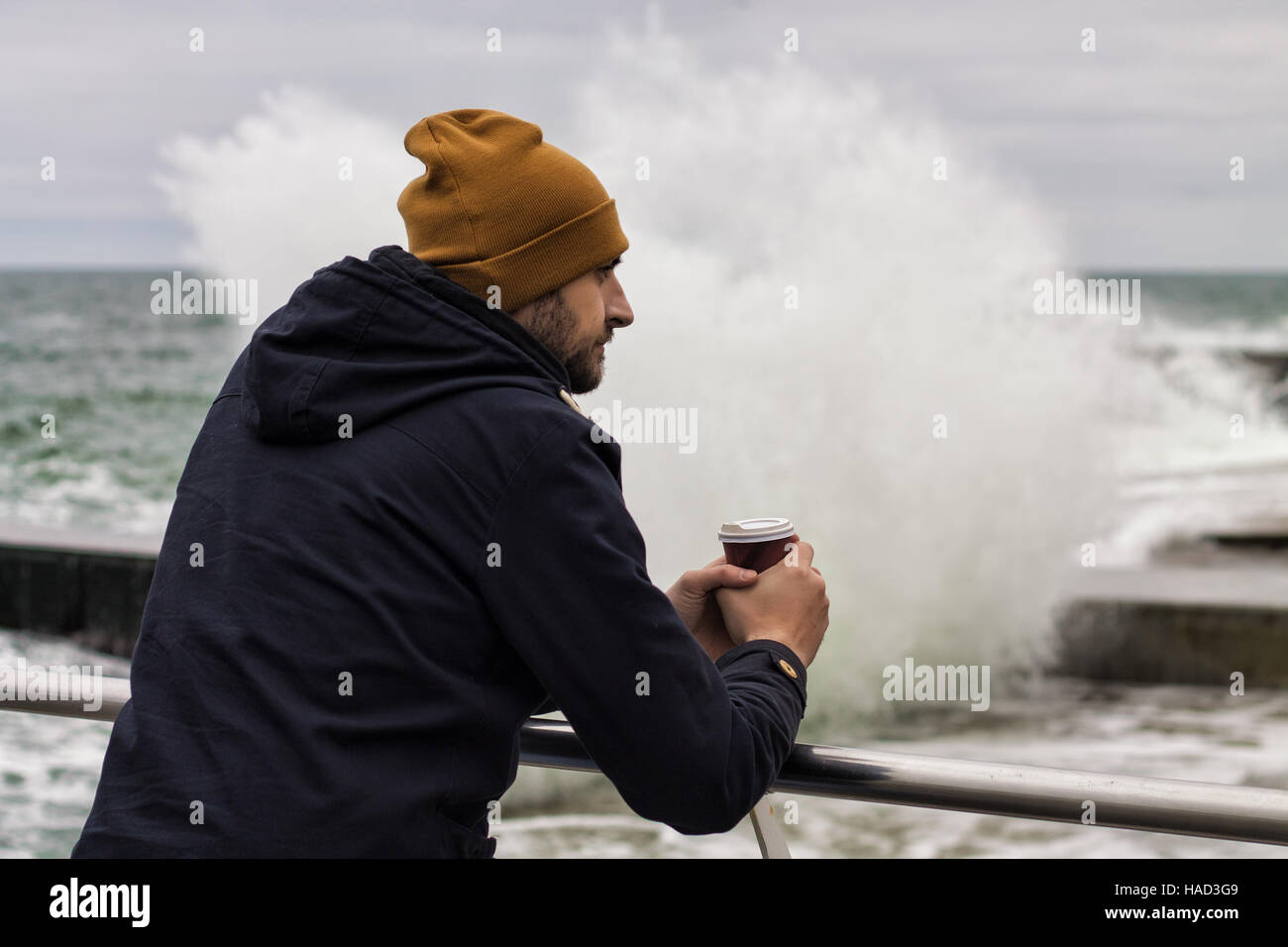 Sad man warms his hands with cup of takeaway coffee. Sea splash is on ...