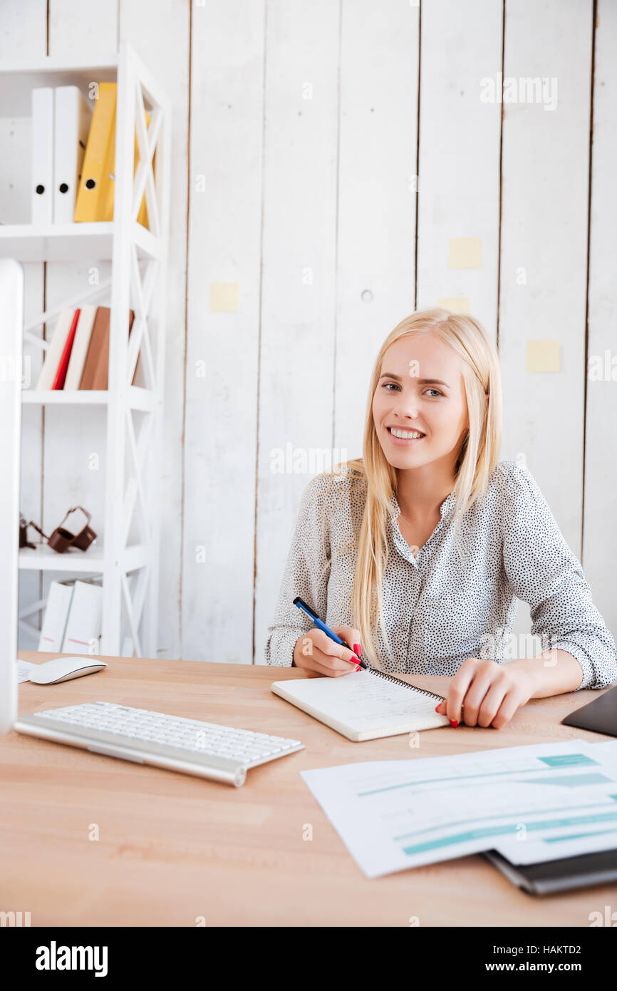 Happy pretty business woman sitting at her workplace and making notes ...