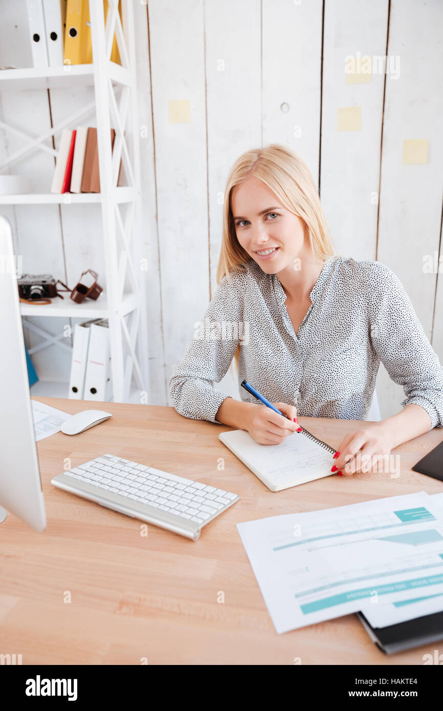 Happy pretty business woman sitting at her workplace and making notes ...