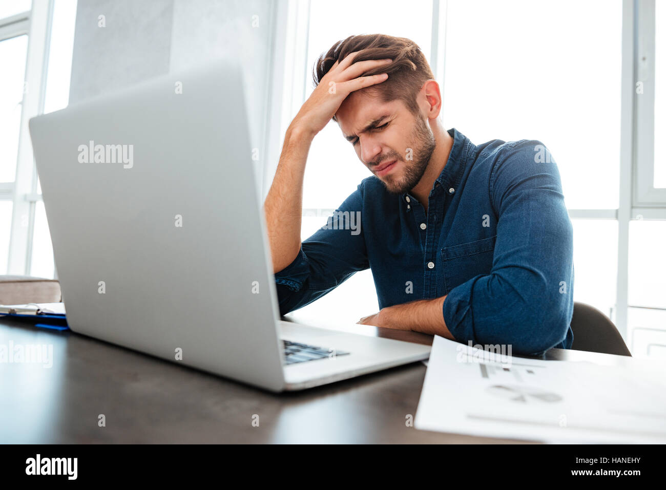 Picture of confused young man sitting near laptop and holding head with ...