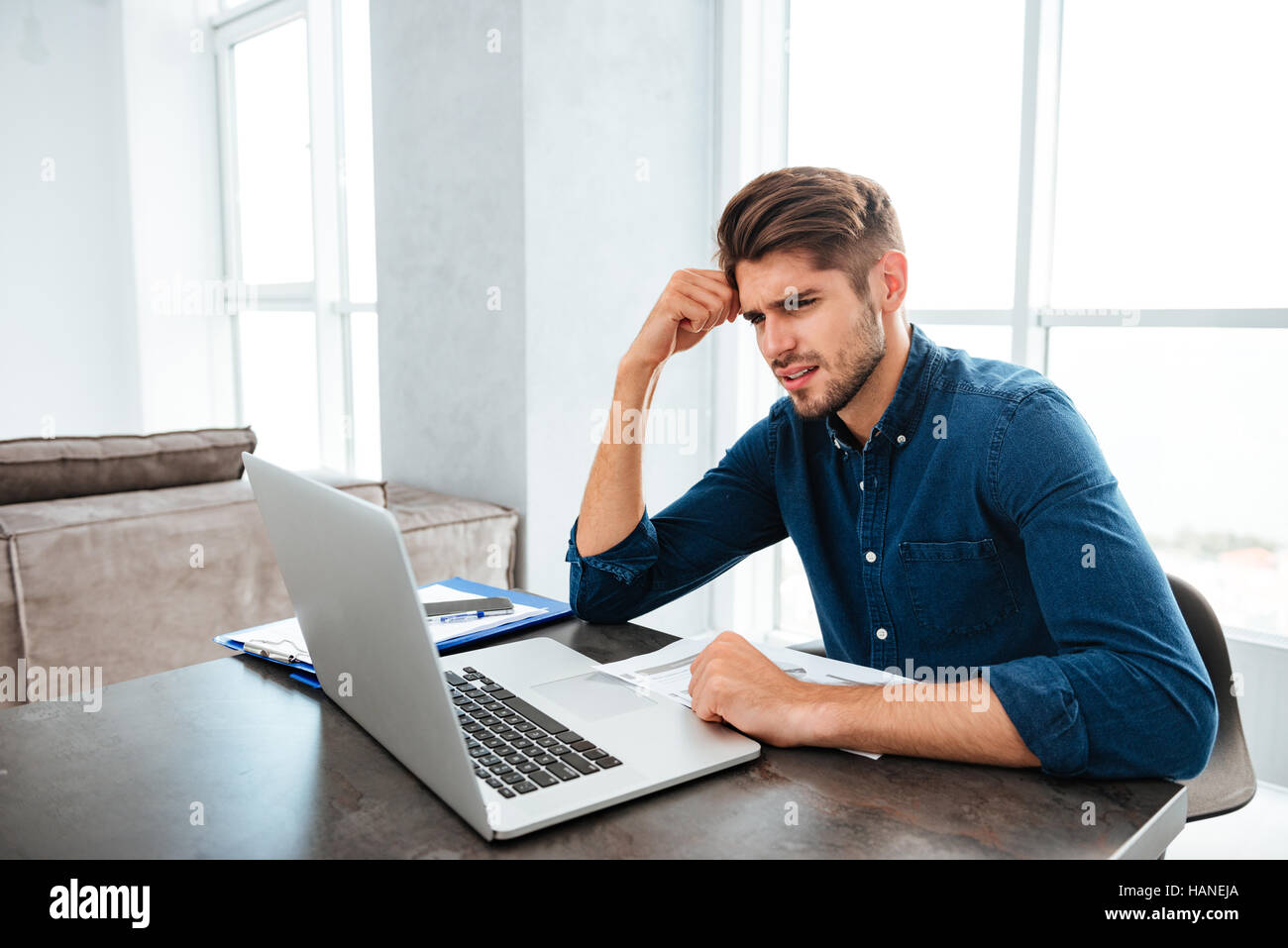Picture of confused young man sitting near laptop and holding head with ...