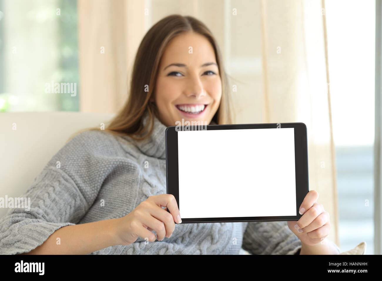 Woman showing a blank tablet screen sitting on a sofa at home in the ...